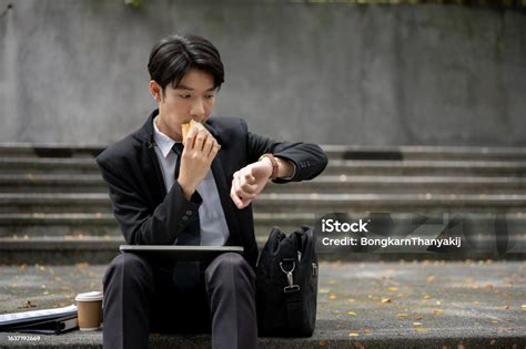 바쁘고 배고픈 아시아 사업가가 시계로 시간을 확인하고 샌드위치를 먹고 있다 먹기에 대한 스톡 사진 및 기타 이미지 먹기