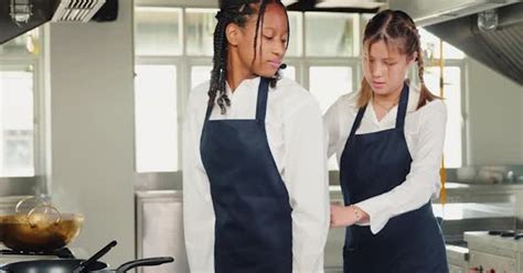 In A Professional Kitchen A Female Chef Assists Her Friend In Tying An Apron Two Women In