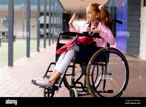 Happy Biracial Schoolgirl In Wheelchair Waving In School Corridor