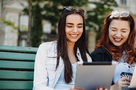 긴 검은 머리를 가진 매력적인 여성의 초상화를 닫고 벤치에 앉아 웃고있는 태블릿을 바라보며 그녀의 여성 친구가 눈을 감고 웃고 있습니다 감정에 대한 스톡 사진 및 기타