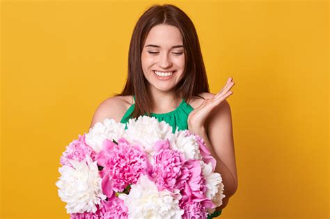 흰색과 장미 모란의 꽃다발을 들고 아름다운 갈색 머리 소녀의 스튜디오 초상화 꽃의 큰 무리와 승리 여자 어두운 머리 여성 녹색 드레스를 입고 노란색 배경 위에 포즈 가정의