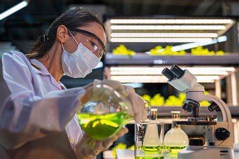 마스크와 장갑을 낀 과학자가 실험실에서 비커에 담긴 녹색 액체를 현미경으로 검사하고 있습니다 실험실 설정은 연구 과학 및 실험에 중점을 둔 밝은 조명입니다 Stem 주제에