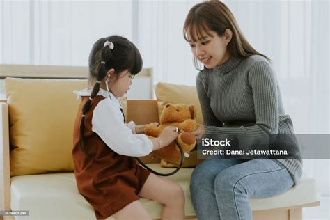 젊은 아시아 어머니와 청진기를 가진 어린 소녀 딸이 소파에 앉아 역할 놀이 의사 곰 인형 장난감을 함께 가지고 있다 20 24