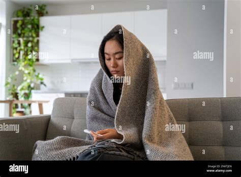Unhealthy Asian Girl Sits Wrapped In Blanket Sadly Looks At Thermometer