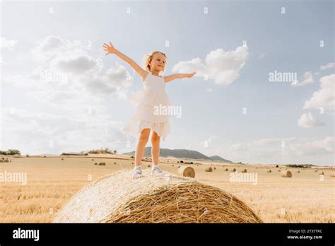 Blond Girl With Arms Outstretched Standing On Bale Of Straw In Stubble