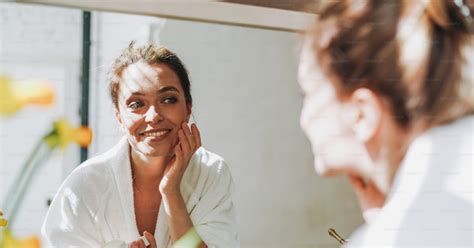 흰색 목욕 가운에 어두운 긴 머리를 가진 젊은 웃는 여자의 아름다움 초상화는 집에서 욕실의 거울 근처에 깨끗하고 신선한 피부 얼굴과 손에 하루 영양 보습 크림을 바르고