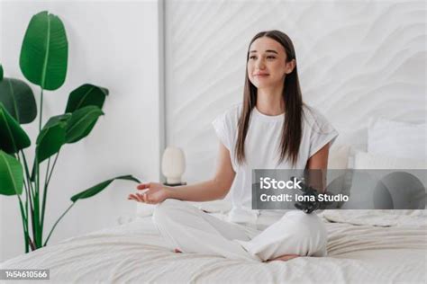흰색 캐주얼 옷을 입은 생체 공학 손 의수를 가진 쾌활한 갈색 머리 백인 소녀는 배경에 큰 식물이 있는 아파트에서 명상 포즈를 취하며 침대에 앉아 있습니다 예쁜 이탈리아