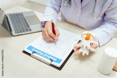 A Male Doctor Wearing Glasses Sits At A Desk Explaining A Spine Model