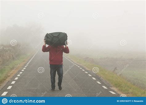 Man In Jeans And Red Mackintosh On Foggy Road With Unrecognisable Suitcase In Tow Royalty Free