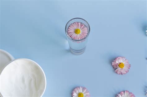 신선한 꽃 데이지는 깨끗한 물 한 잔과 파란색 배경에 크림 화장품이있는 흰색 항아리가있는 구석에 배치되어 있습니다 0명에 대한