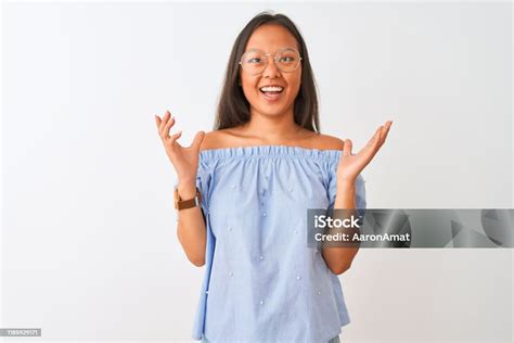 파란색 티셔츠와 안경을 쓰고 고립된 흰색 배경에 미친 듯이 미친 듯이 외치며 공격적인 표정과 팔을 들고 고함을 지르는 젊은 중국