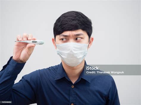 마스크를 쓰고 있는 아시아 남자 심각한 얼굴이 그의 손에 하얀 배경에 온도계를 바라보고 있다 30 34세에 대한 스톡 사진 및