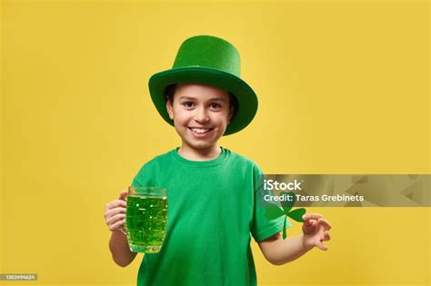 레프레콘 아이리쉬 모자를 쓴 어린 소년은 녹색 음료를 입은 유리와 클로버 잎미소를 들고 카메라를 들고 포즈를 취하고 있다 세인트 패트릭의 날 3월에 대한 스톡 사진 및 기타