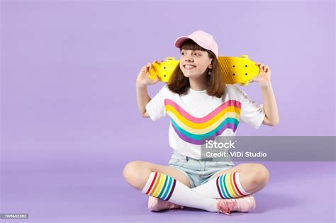 선명한 옷을 입고 예쁜 십 대 소녀 스튜디오에서 보라색 파스텔 벽 배경에 고립 된 옆에 노란 스케이트 보드를 들고 앉아 사람들은 진심으로 감정 라이프 스타일 개념입니다 복사