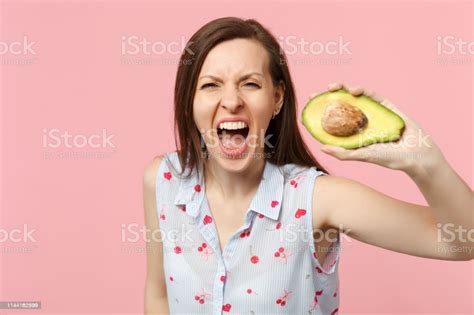 야생 분홍색 파스텔 벽 배경에 고립 된 신선한 잘 익은 녹색 아보카도 과일의 절반을 들고 여름 옷에 젊은 여자를 비명 사람들은