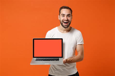 밝은 오렌지 색 배경 스튜디오 초상화에 고립 포즈 캐주얼 흰색 티셔츠에 흥분 젊은 남자 사람들 라이프 스타일 개념 복사 공간을 모의합니다 빈 빈 화면으로 노트북 Pc