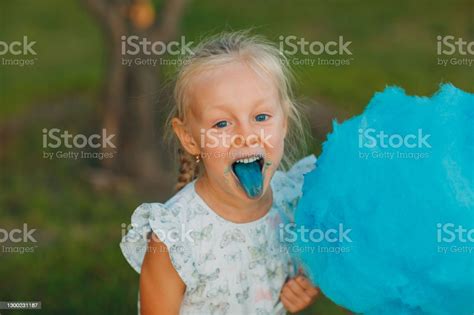 Little Blond Girl Eating Cotton Candy And Shows Blue Tongue In The Park