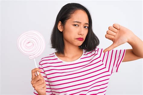 화가 얼굴 엄지 손가락으로 싫어하는 것을 보여주는 부정적인 기호 거부 개념으로 고립 된 흰색 배경 위에 서있는 롤리팝을 먹는 젊은 아름다운 중국 여성 갈색 머리에 대한 스톡