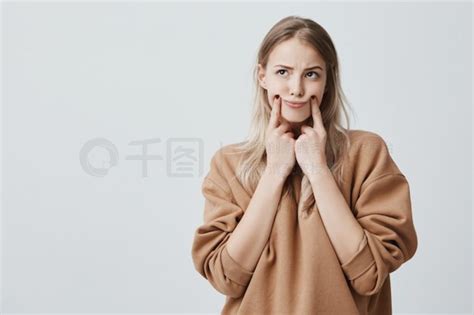 不悦高清图片 漂亮的金发女人做鬼脸，用手指抚摸她的脸颊，向上看，表情不悦。面部表情和负面情绪 产品实拍免费下载格式编号