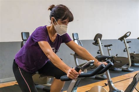 새로운 정상 사회적 거리 Covi19 코로나 바이러스의 낮은 용량으로 인해 빈 체육관에서 고정 자전거에 얼굴 마스크를 가진 젊은 백인 소녀 보호 마스크에 대한 스톡 사진 및