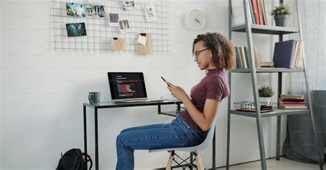 캐주얼웨어와 이어폰을 입은 쾌활한 젊은 혼혈 여학생이 스마트폰으로 스크롤하는 동안 노트북 앞 작은 테이블 옆에 앉아 있다 사진 Unsplash의 기술 이미지