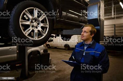 자동차 정비사 제복을 입은 기술자가 보증 차량 유지 보수 및 서비스 중에 수리소의 호이스트에서 들어 올려진 차량을 수리하기 위한 클립보드의 체크리스트를 제작합니다 개선에