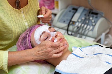 숙련 된 미용 학자는 얼굴 클렌징 및 미용 치료 중에 소파에 놓여 있는 어린 소녀의 얼굴에 흰 크림 마스크를 적용 합니다 내각 미용사 피부 건강과 청소년 거실에 대한 스톡