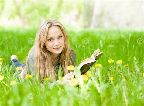 Девушка лежа на траве с одуванчиками читая книгу и смотря камеру