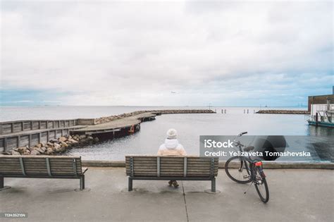 젊은 백인 여자는 흐린 날씨에 겨울에 코펜하겐 덴마크의 바닷가에 발트 해를 내려다 볼 수 있는 나무 벤치에 그녀의 뒤에 앉아 인근에 있는 소녀 산책 곤 오스카 자전거 가을에