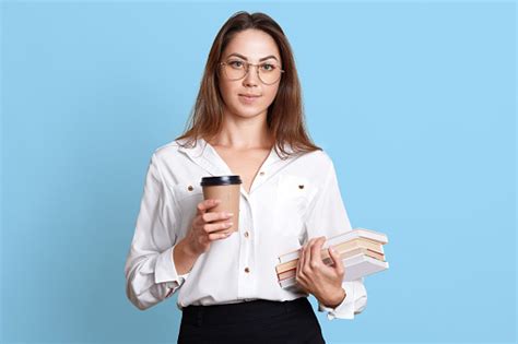 유행 안경에 하드 작업 심각한 젊은 여자의 초상화 파란색 배경 위에 고립 유럽의 갈색 머리 모델은 책의 무리와 커피의 종이 컵을 보유 하 고 있습니다 광고 공간을 복사