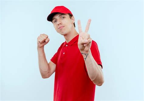 빨간 제복을 입은 젊은 배달 남자와 흰 벽 위에 자신감이 서있는 승리 기호를 보여주는 주먹 떨림 모자 무료 사진