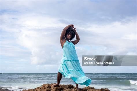 해변의 바위 위에 파란 옷을 입고 팔을 흔드는 검은 파워 머리를 한 아름다운 여자 배경 하늘과 바다에서 30 34세에 대한 스톡