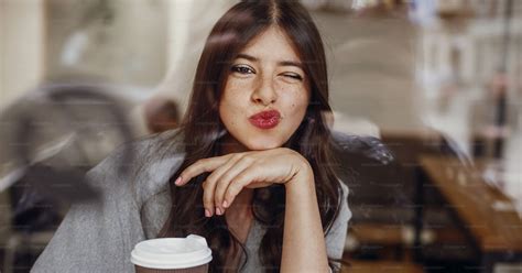 키스를 보여주는 화려한 젊은 여성 커피 한 잔과 함께 카페에 앉아 도시 거리의 창문을 통해 바라보고 있습니다 아름 다운 머리 키스와 미소를 가진 세련 된 힙 스터 소녀