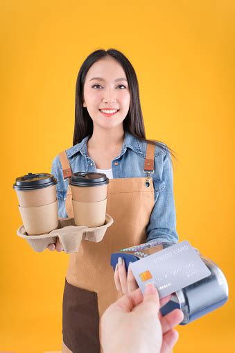 예쁜 아시아 여성 스마일 커피 프리랜서 자신의 커피 바 숍은 커피 종이 컵을 들고 자신의 상점 노란색 배경에서 신용 카드를 스 와이프하는 단말기 돈을 통해 신용 카드로