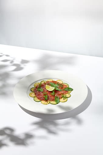 햇빛 그림자가있는 흰색 테이블에 호박과 오이를 곁들인 쇠고기 카르 파치오 고기의 전채 여름 식사에 쇠고기 Carpaccio 이탈리아 스타일로 얇게 썬 생고기 미적 메뉴 카르