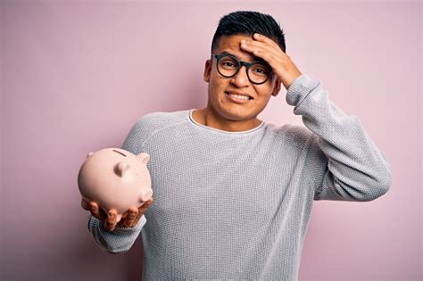 고립 된 분홍색 배경 위에 돼지 저금통을 들고 있는 젊은 잘생긴 라틴 남자는 머리에 손으로 스트레스를 받고 수치심과 깜짝 놀란