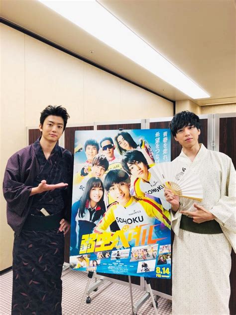Two Men Standing Next To Each Other In Front Of A Poster