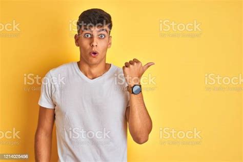 흰색 티셔츠를 입은 젊은 인도 남자가 고립 된 노란색 배경 위에 서 놀란 손으로 손가락을 옆으로 가리키고 입을 벌리고 놀란 표정