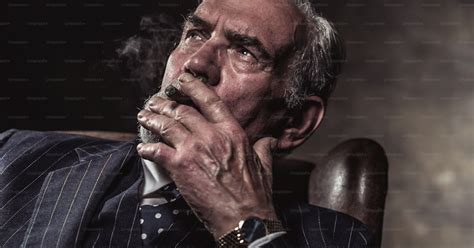 의자에 앉아 있는 특징적인 고위 사업가 시가를 피우고 있다 회색 머리와 턱수염은 파란색 줄무늬 양복과 넥타이를 착용하고 있습니다 갈색 벽에 대하여 사진