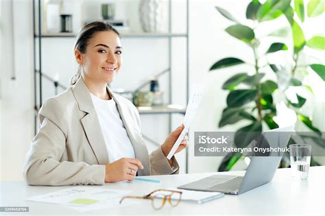 아름답고 성공한 백인 사업가 사무실에 앉아서 작업용 책상에서 일하는 동안 그래프와 차트가 있는 보고서를 들고 카메라를 바라보며 미소 짓고 있습니다 35 39세에 대한 스톡