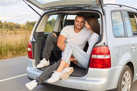 Portrait Of Loving Couple Sitting In Car Trunk Wearing White Shirts