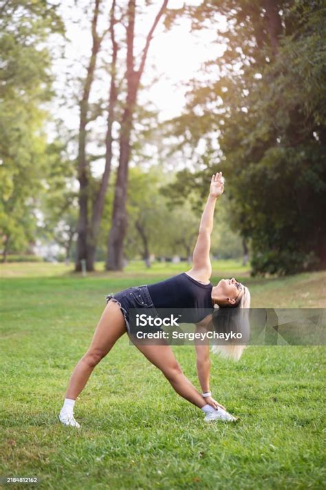 햇볕이 잘 드는 따뜻한 저녁에 공원에서 짧은 반바지와 탱크 탑을 연습하는 Trikonasana 운동 삼각형 포즈를하는 유럽 외모의 스포티 한 여성 가냘픈에 대한 스톡 사진