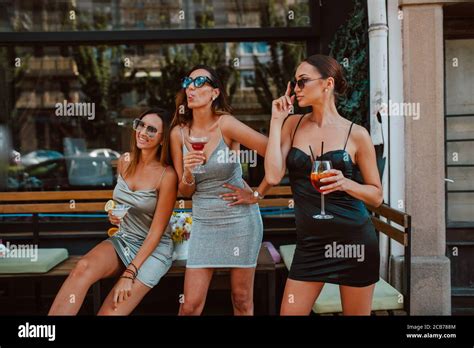 Three Attractive Caucasian Women In Short Tight Dresses Are Drinking Cocktails In A Cafe Garden