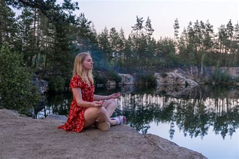 소나무 숲의 침수 된 화강암 채석장의 배경에 절벽가장자리에 앉아 일몰에 명상 빨간 드레스를 입은 젊은 금발 의 여자 자연과의 통일 화재의 연기 Mental Wellbeing