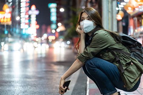 아시아 의 젊은 여성 배낭 가면을 쓰고 밤에 도시에서 버스를 기다립니다 아름다운 관광 여행자 소녀는 다채로운 빛으로 거리에서 혼자 여행하고 전염병 동안 휴가 휴가를