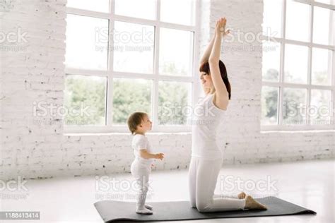 흰색 옷을 입은 스포츠 젊은 어머니는 큰 창문이있는 가벼운 체육관에서 아기 소녀와 함께 피트니스와 요가에 종사합니다 2명에 대한