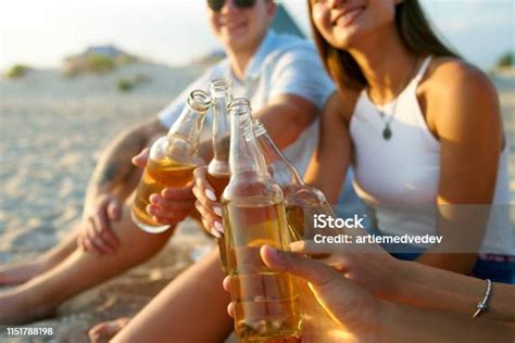 상쾌한 음료를 즐기고 석양에 해변에서 휴식을 즐기는 친구 들의 그룹 젊은 남성과 여성은 따뜻한 여름 저녁에 모래에 앉아 맥주를