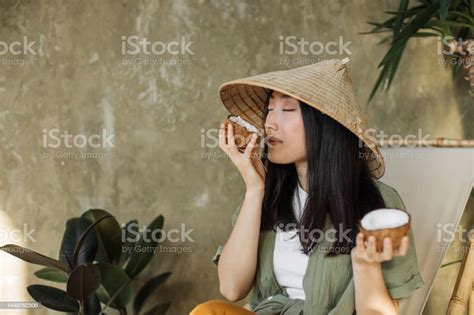 신선한 코코넛 반쪽의 냄새를 즐기는 전통적인 원뿔형 모자를 쓴 매력적인 아시아 여성 25 29세에 대한 스톡 사진 및 기타