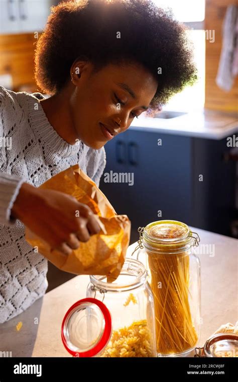 Happy african american woman pouring pasta from paper bag to storage jar in sunny kitchen Stock ...