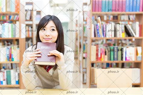 عکس دختر در کتابخانه نشسته پشت میز با یک کتاب در دست عکس با کیفیت و